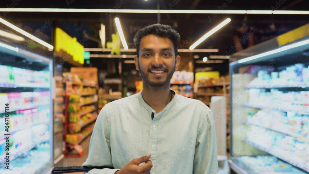 Smiling indian man walking in aisle with shopping basket at supermarket ...