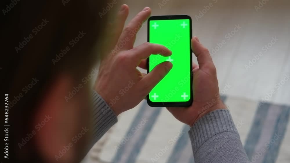 Young man sitting at home holding smartphone greenscreen mock-up in hand. Male person using ...