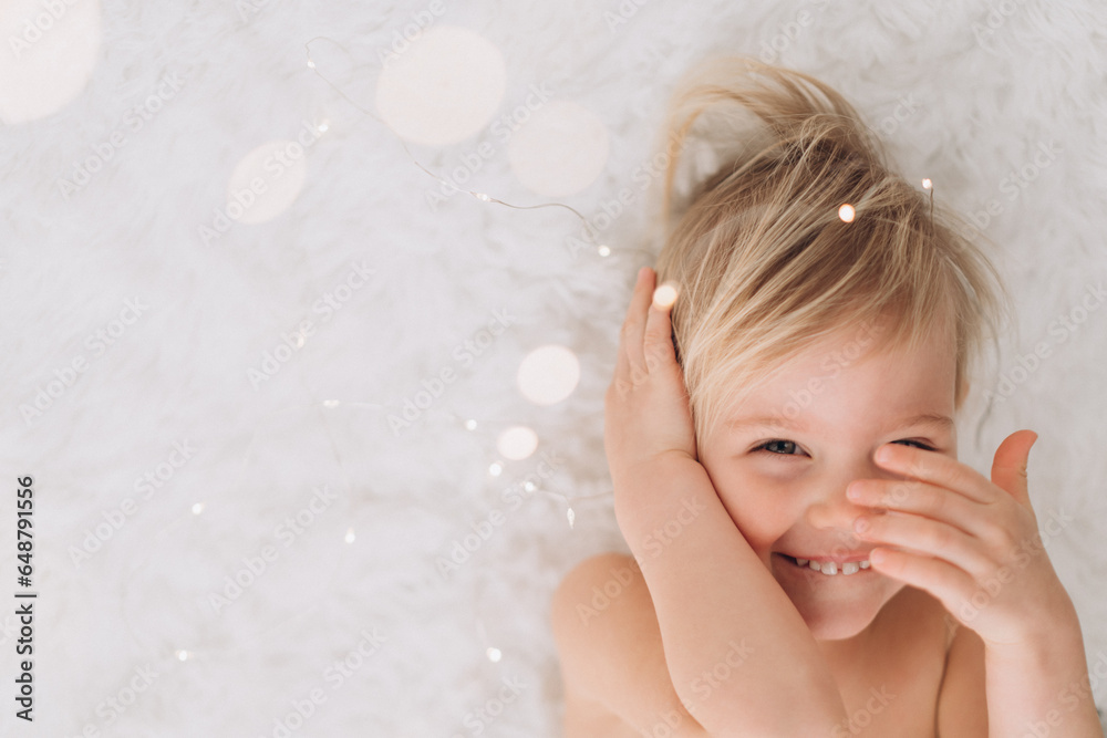 A naked child in the lights from a garland closes face with his hands ...