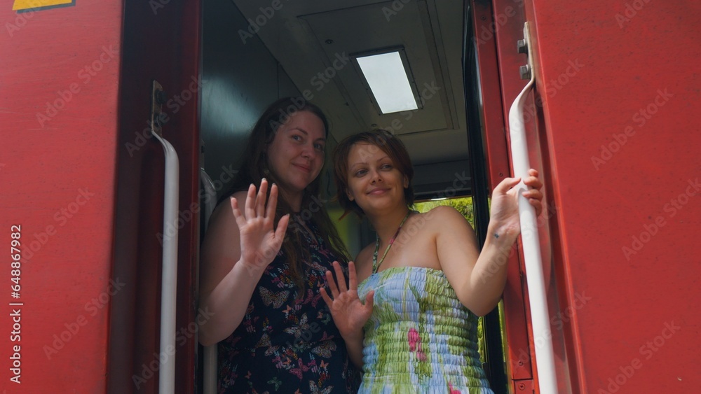 Two girls lean out of train vestibule and wave to people on platform ...