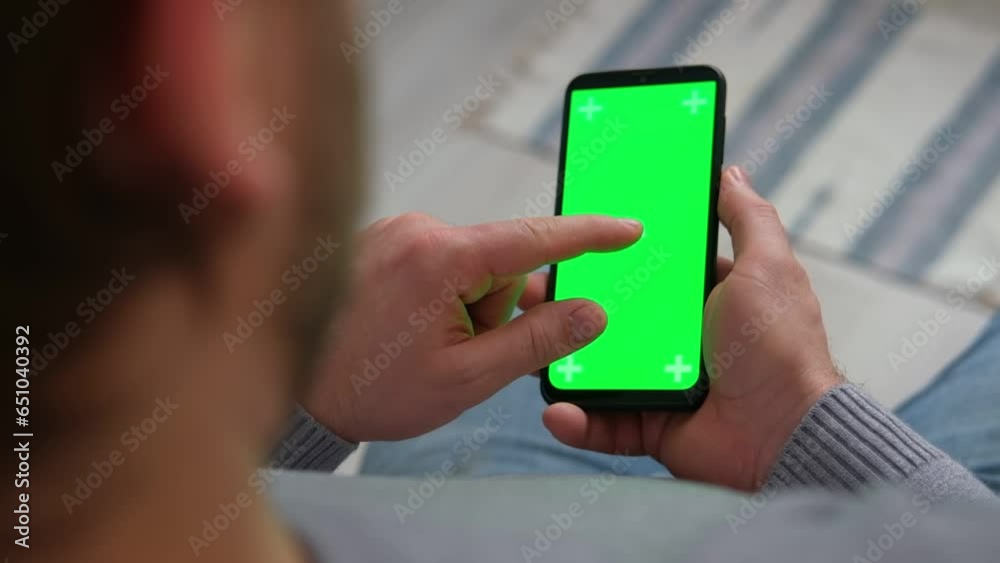 Young man sitting at home holding smartphone greenscreen mock-up in hand. Male person using ...