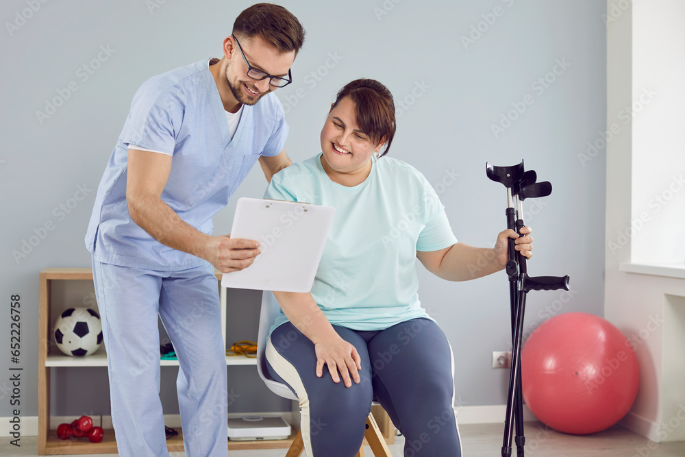 Cheerful friendly nurse showing fat young patient with crutches a ...