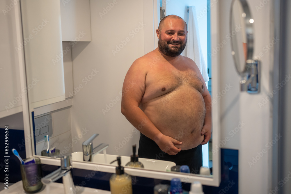 Fat obese man smiling looking in mirror. Bald bearded overweight guy in ...