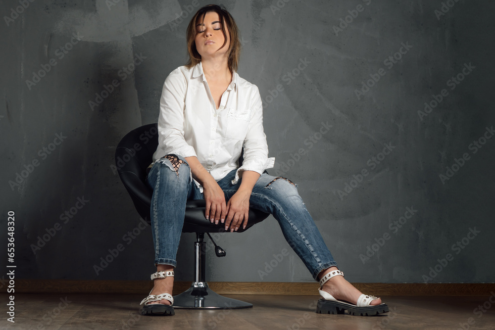 Wonderful lady with closed eyes in shirt and jeans sitting on chair ...