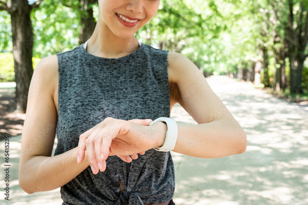 Portrait of an Asian woman jogging and using a smartwatch in a park ...