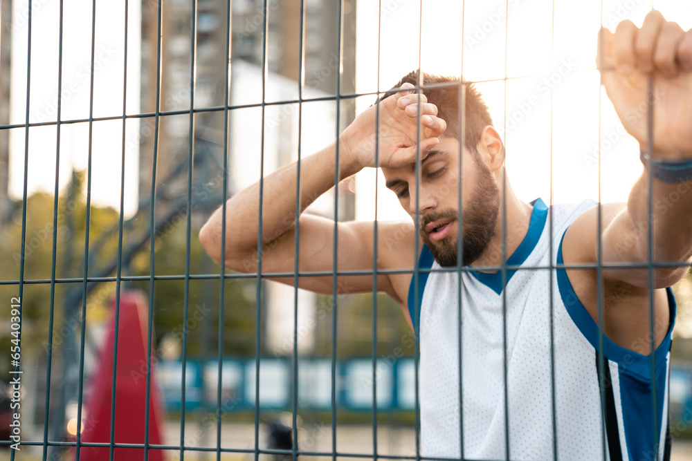 Handsome exhausted male basketball player leaning on fence while taking ...