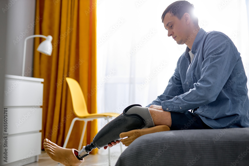 man wearing artificial leg sitting on bed at home, preparing prosthesis ...