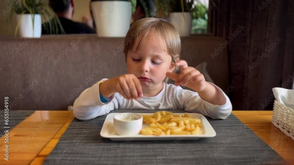 Cute little boy eating french fries dips potatoes in white sauce, puts ...