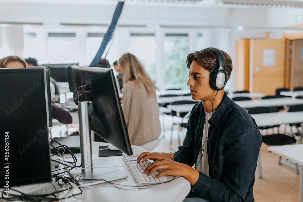 Teenage boy listening through headphones while using computer in school ...