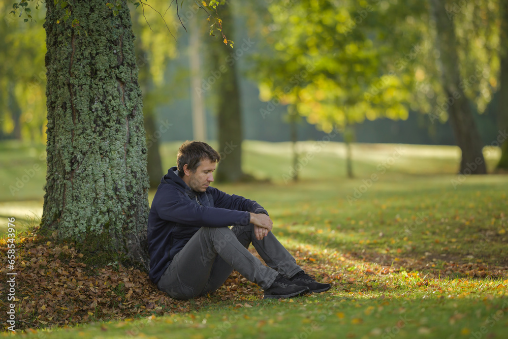 Sad young adult man sitting alone on grass at tree in autumn day ...