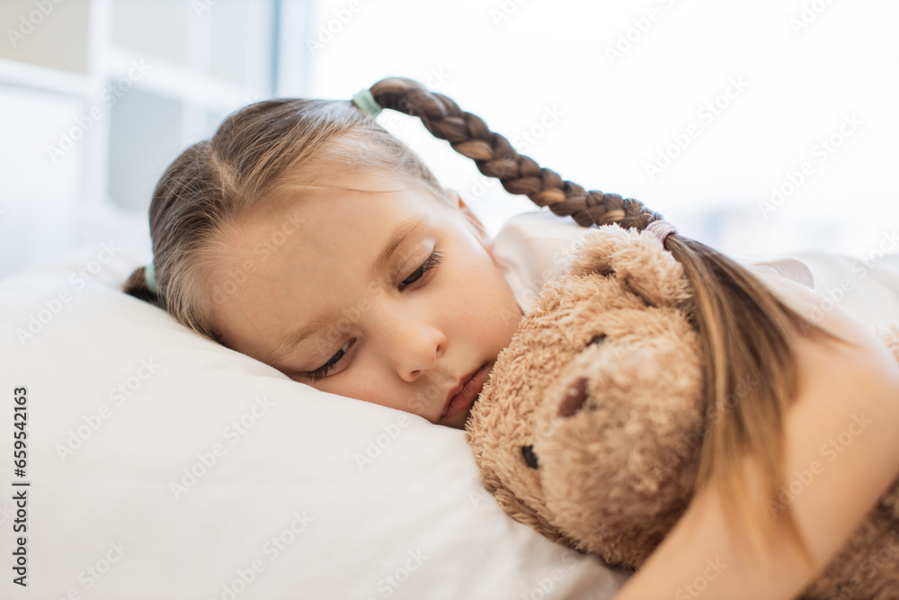 Caucasian little girl with gloomy look sleeping in white bed and seeking solace in embrace of ...