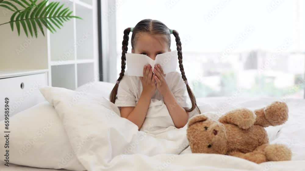 Sad caucasian girl sitting on bed with her teddy bear toy and blowing nose in white napkin ...