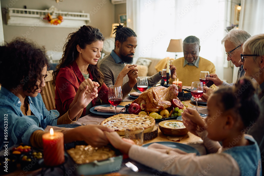 Multiracial extended family holding hands and praying while gathering for Thanksgiving meal at ...