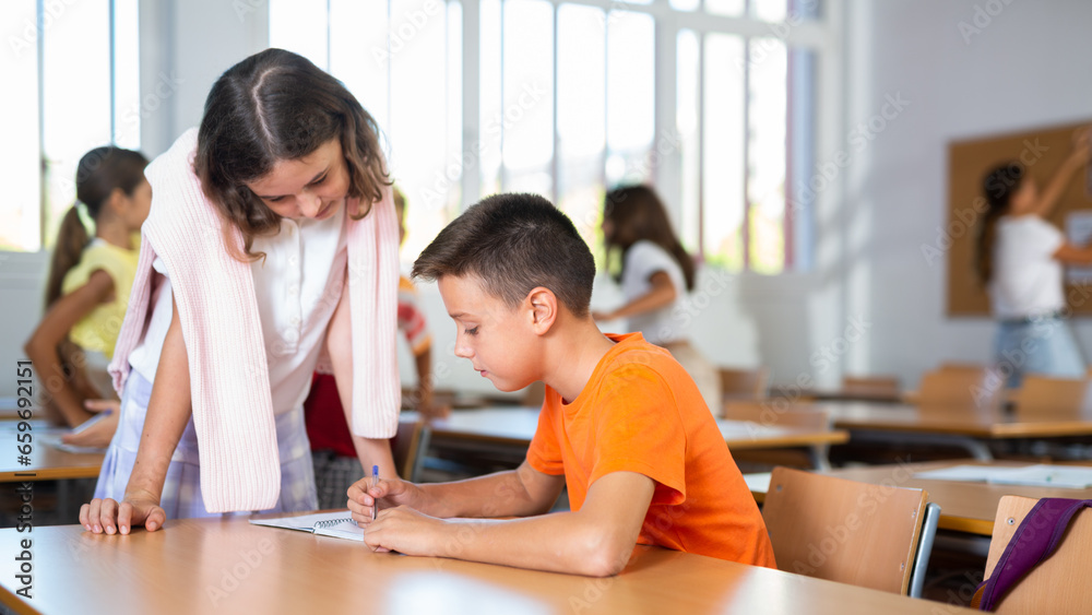 Positive friendly tween girl helping classmate sitting at school desk ...