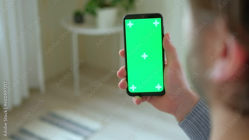 Young man sitting at home holding smartphone greenscreen mock-up in hand. Male person using ...