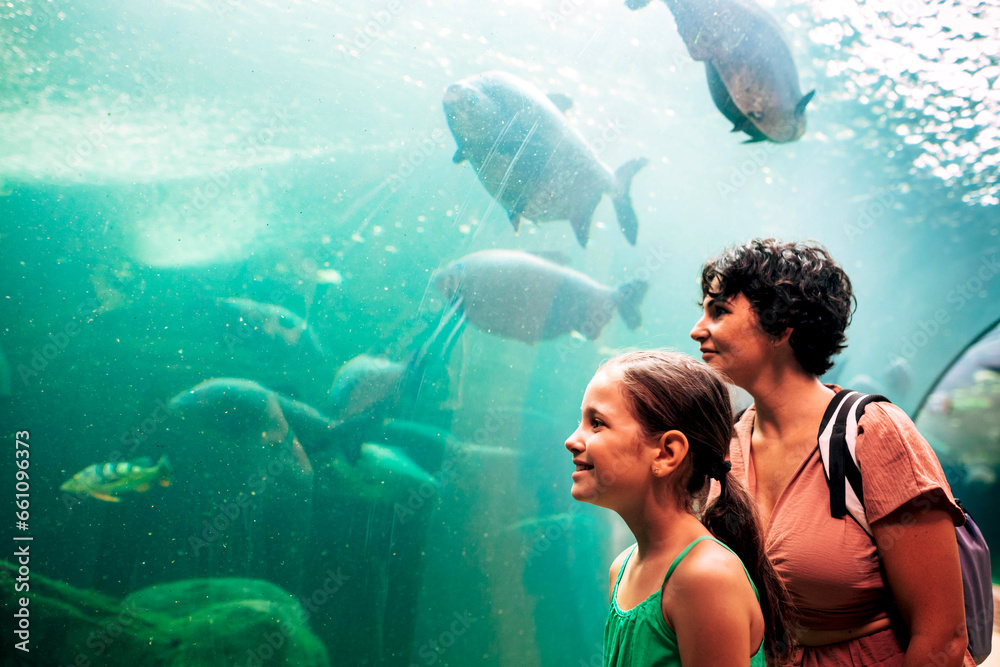 Smiling mother and daughter standing near aquarium glass wall with ...