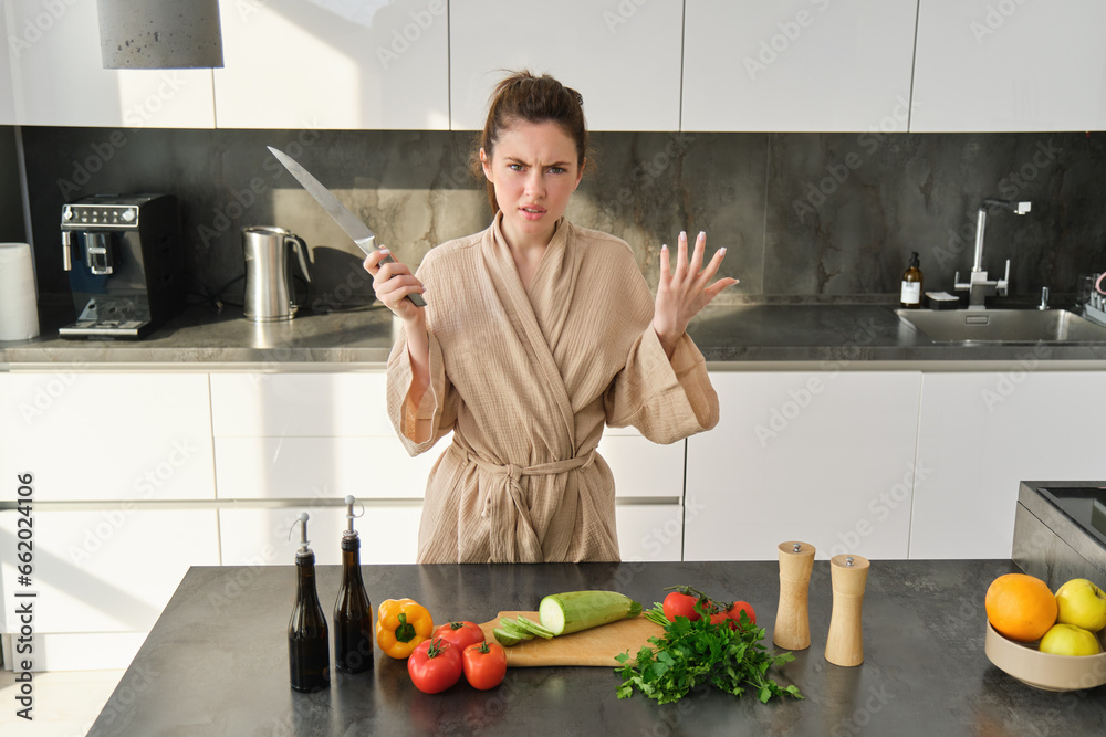 Portrait of annoyed woman with knife, angry while cooking in the ...