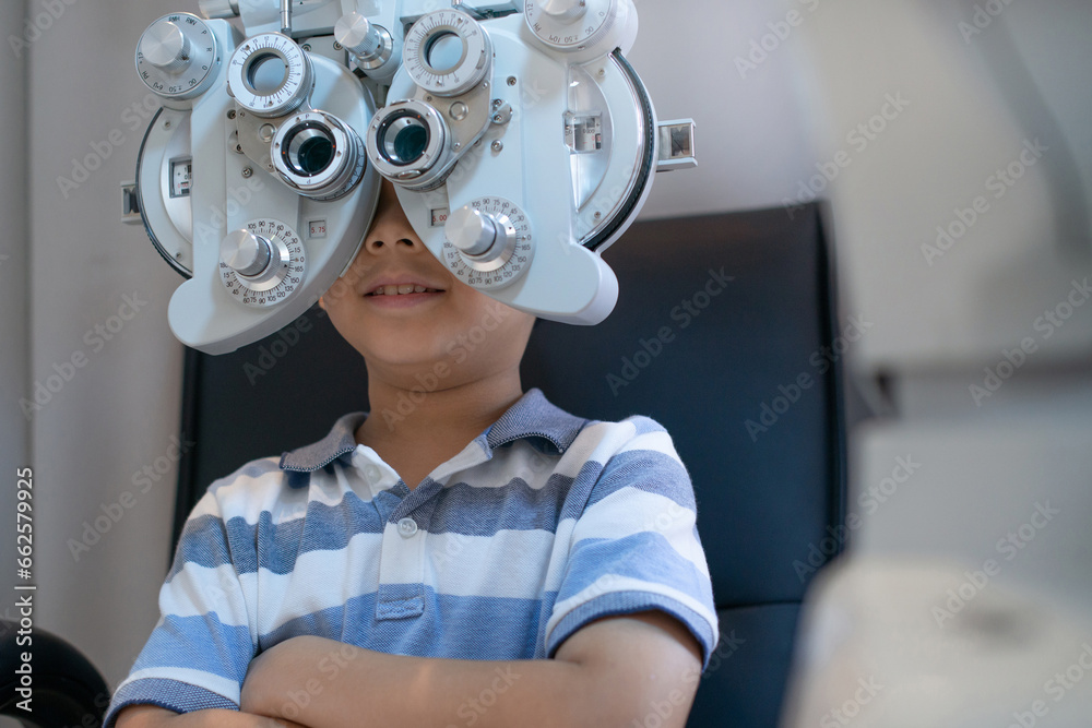 Asian boy crossing his arms and looking through optical phoropter ...