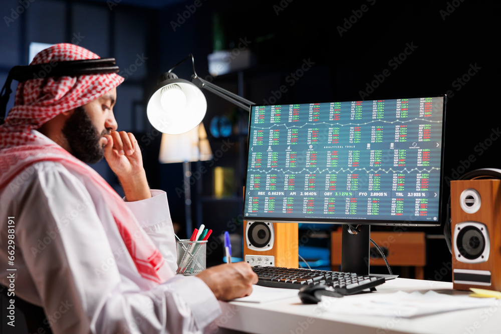 Middle Eastern man seated on his desk carefully researching and jotting ...