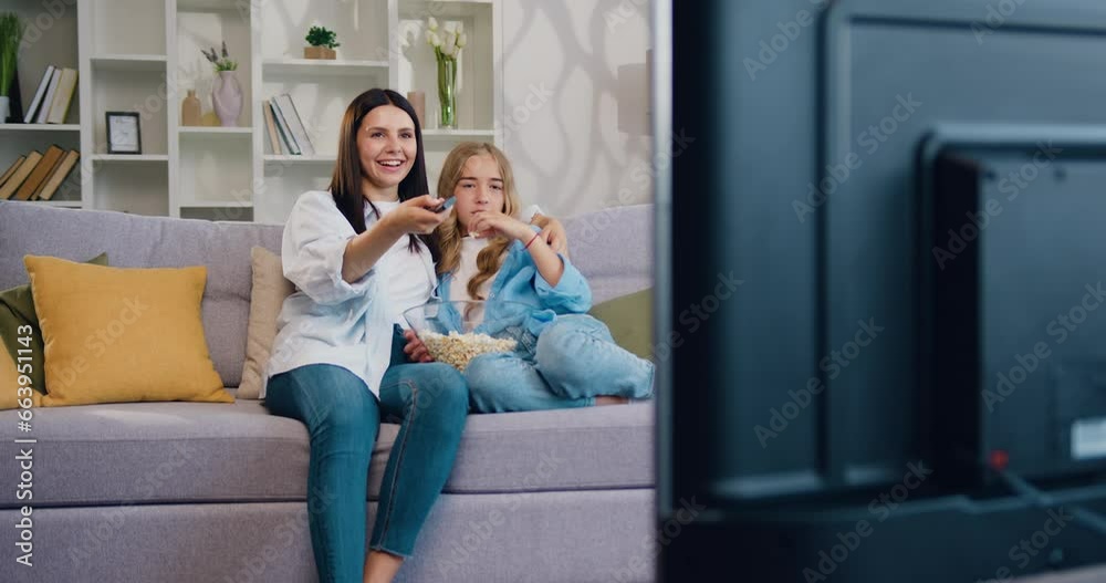 Cute teen daughter and smiling mother laughing sitting on couch eating popcorn enjoy funny ...