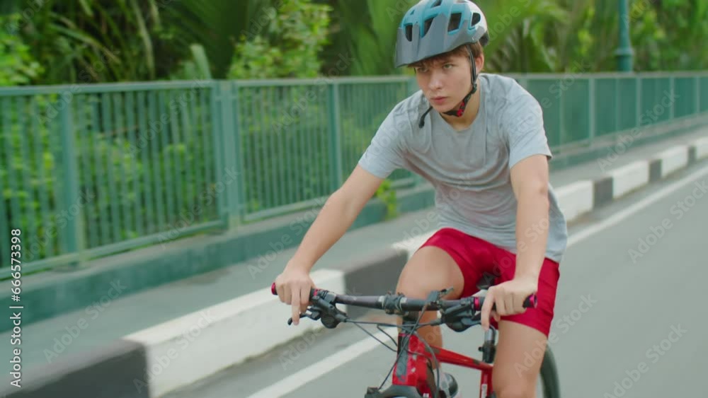 Teen boy in sportswear and protective helmet riding bicycle along road outdoor on summer day ...