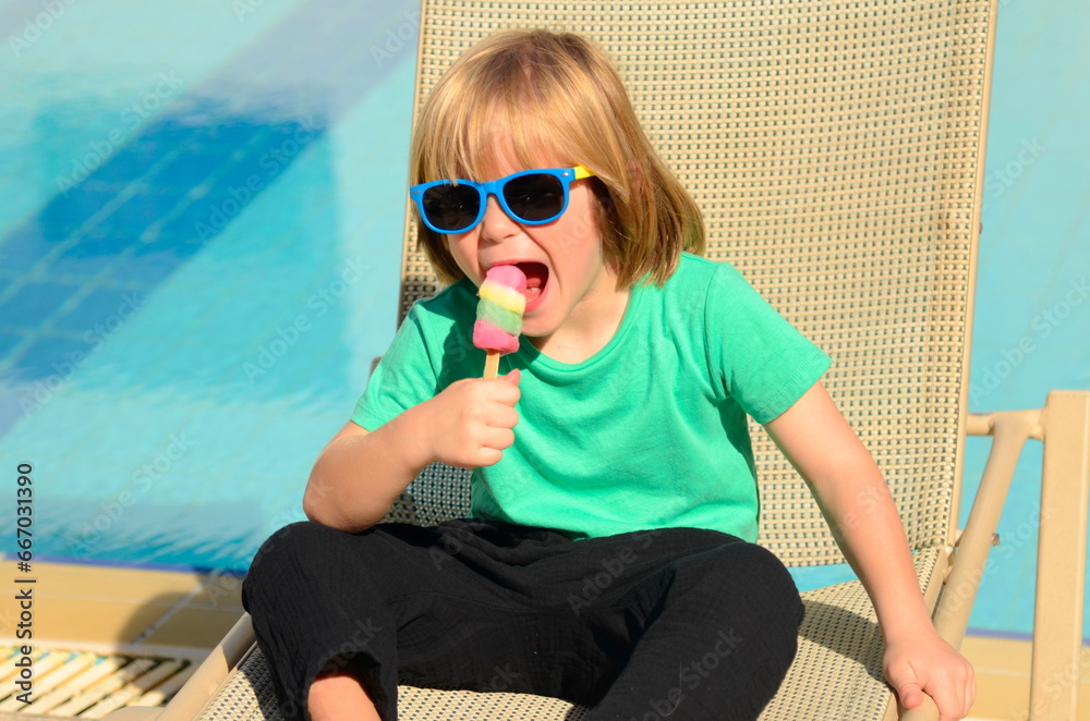 Positive kid lies in a sun lounger and eats ice cream near the pool ...