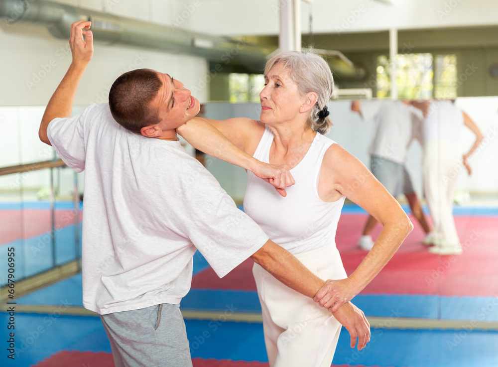 Focused elderly woman performing elbow strike and wristlock, painful control move to immobilize ...