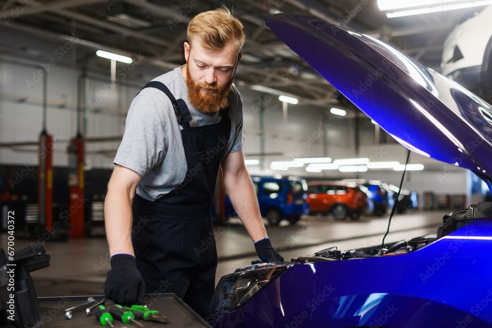 A red-bearded male car mechanic in uniform takes tools from the table ...