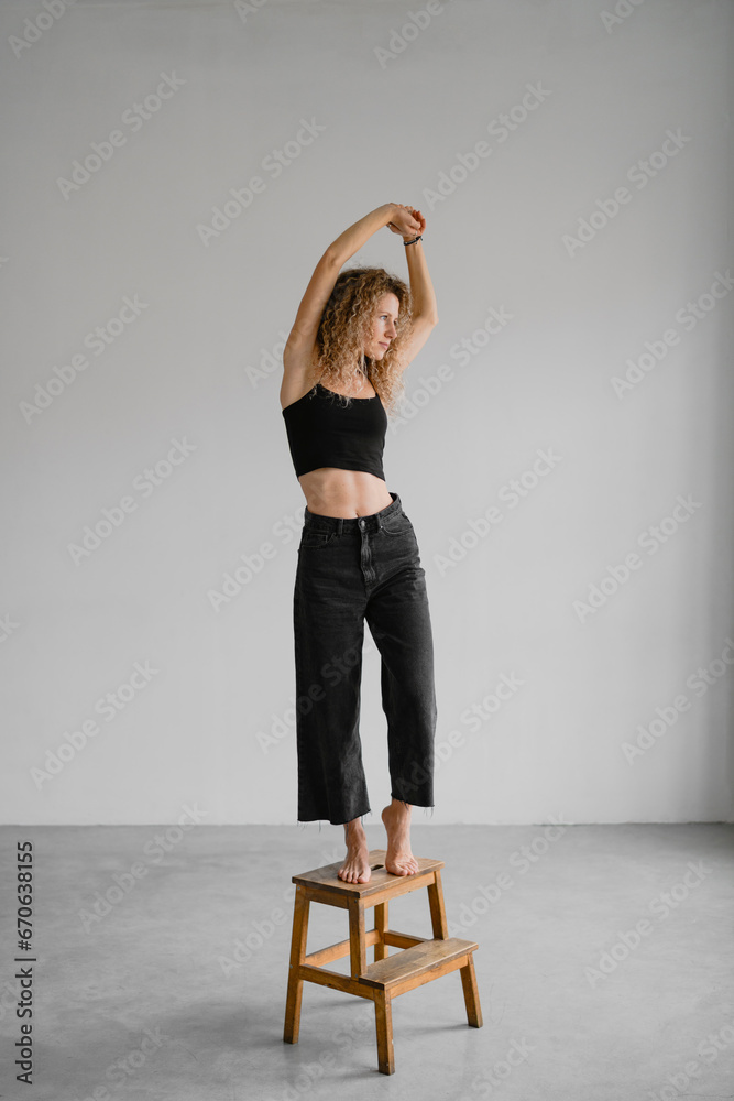 Barefoot woman in dark jeans and top posing on step stool on grey ...