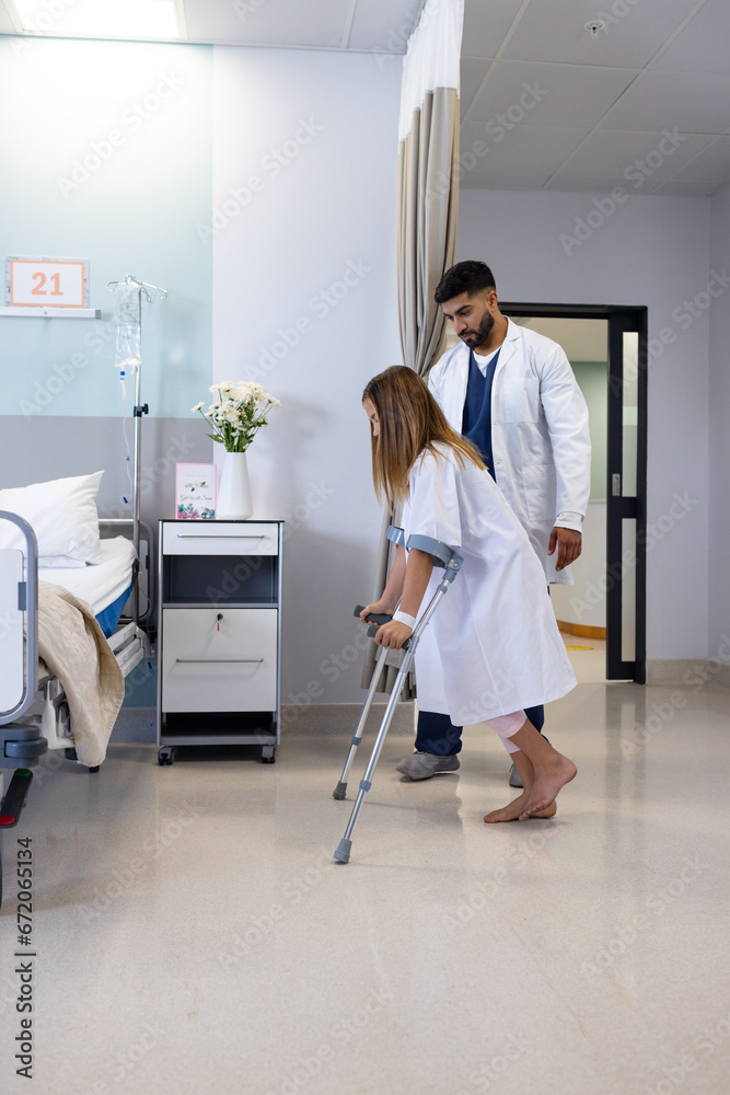 Diverse male doctor observing girl patient use crutches in hospital ...