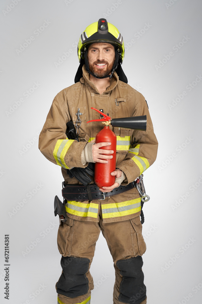 Smiling male firefighter in full gear holding fire extinguisher indoor ...