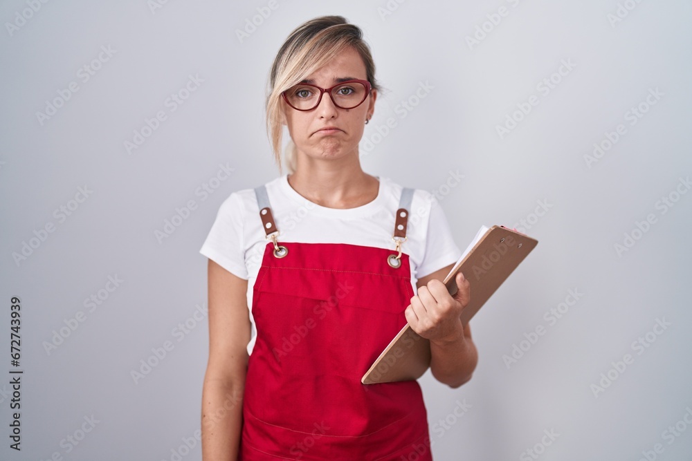 Young blonde woman wearing waiter uniform holding clipboard depressed ...