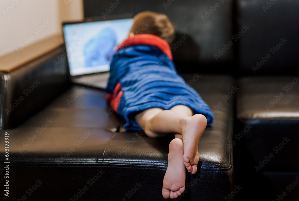 A boy child in pajamas lies, rests on a black leather sofa, watching a ...