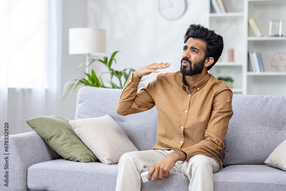 Young Indian man with beard sits in the living room in summer, fanning ...