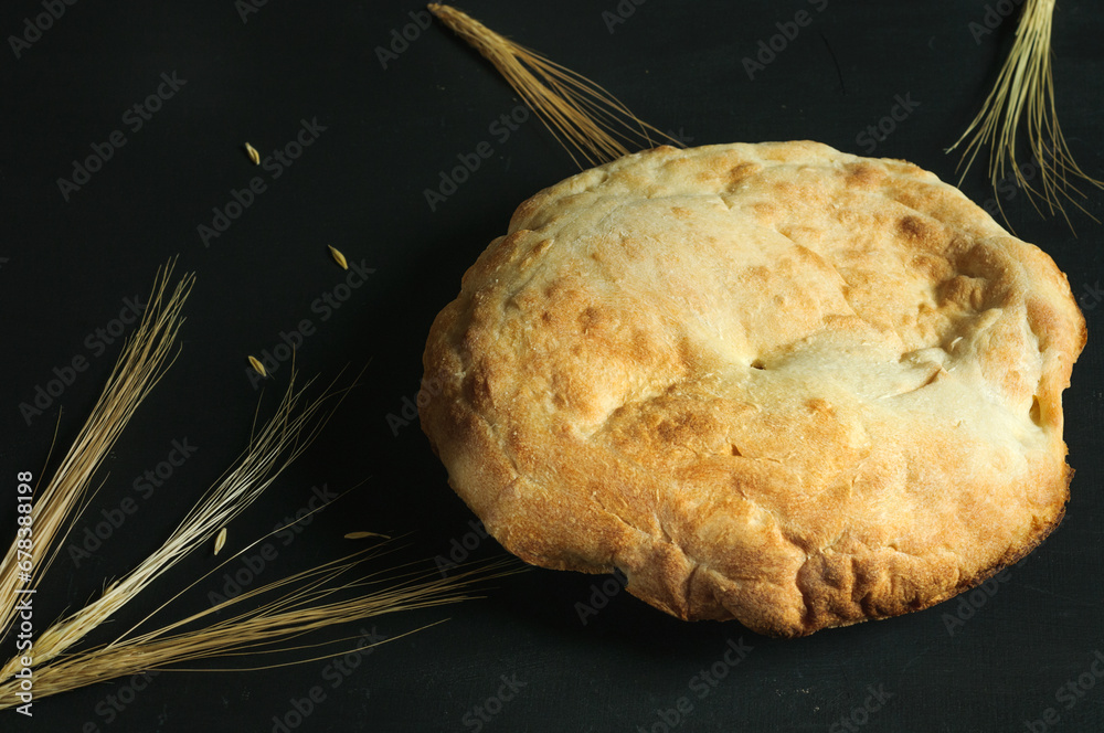 Round flat bread on a black background with ears of rye. Natural light ...