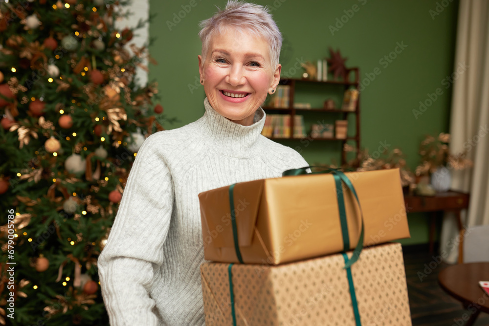 Cheerful granny in sweater carrying big gift boxes trying to hide them ...