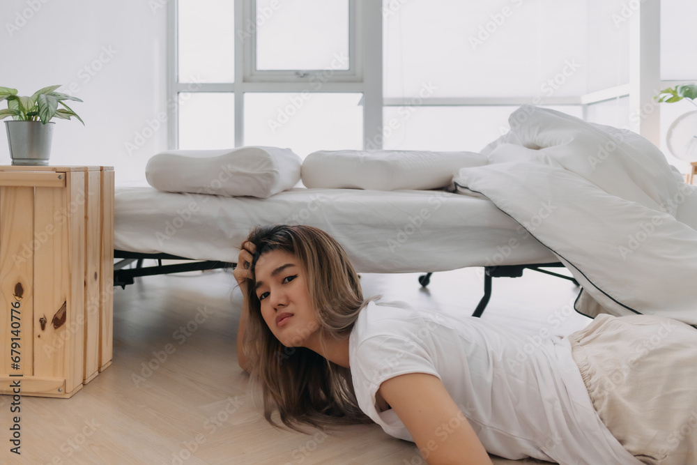 Asian Thai woman wake up from falling down from white bed, lying on floor with funny drowsy and ...