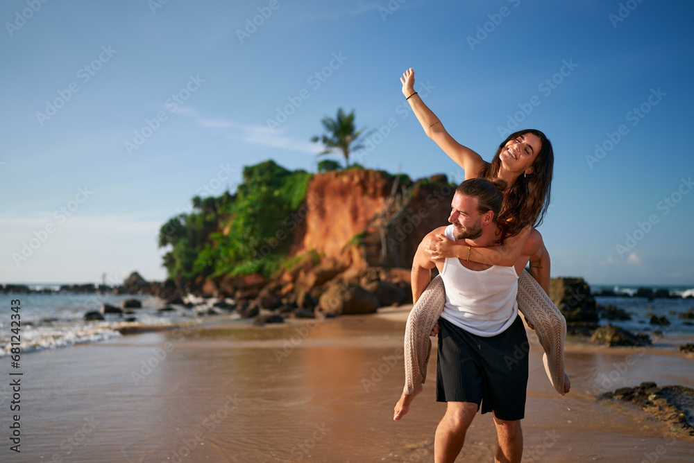 Joyful couple enjoys piggyback ride on tropical beach, woman raises ...
