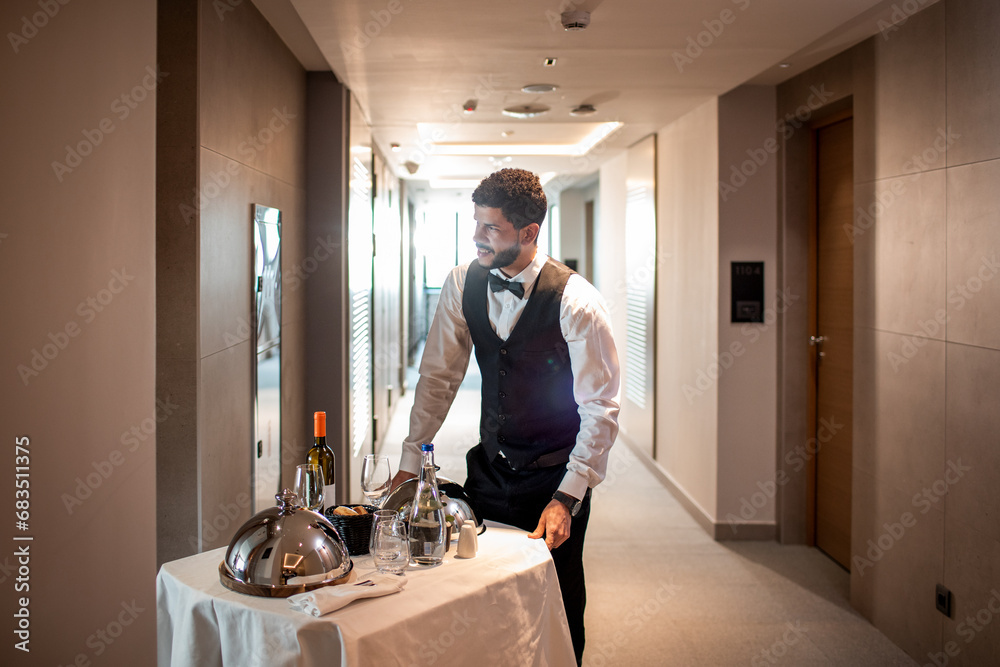Young waiter delivering tray with food in a room of hotel doing room ...
