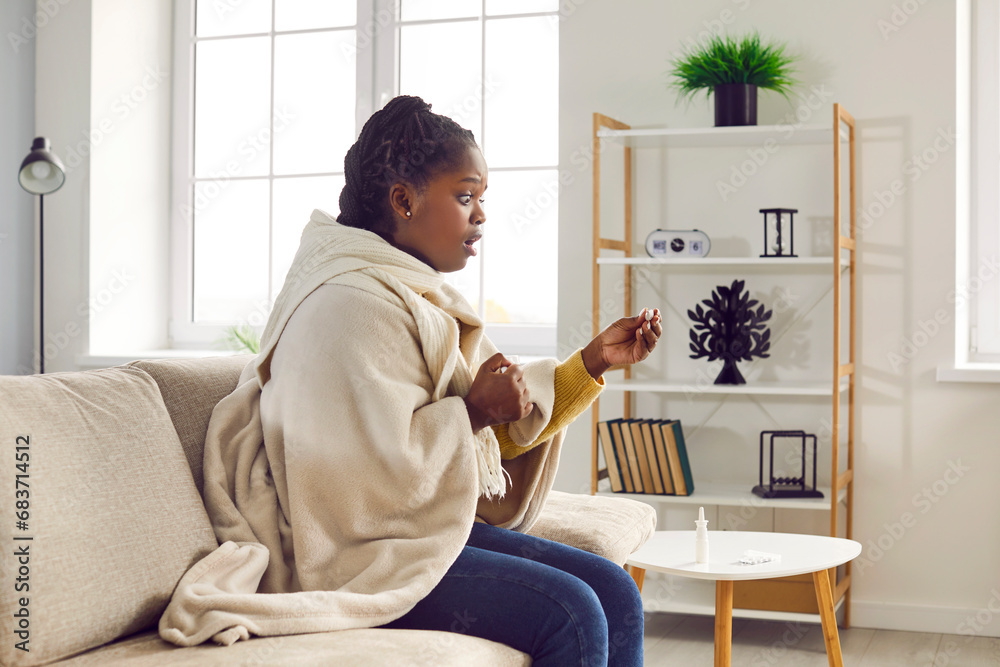 Sick young African American woman with fever sitting on sofa at home ...