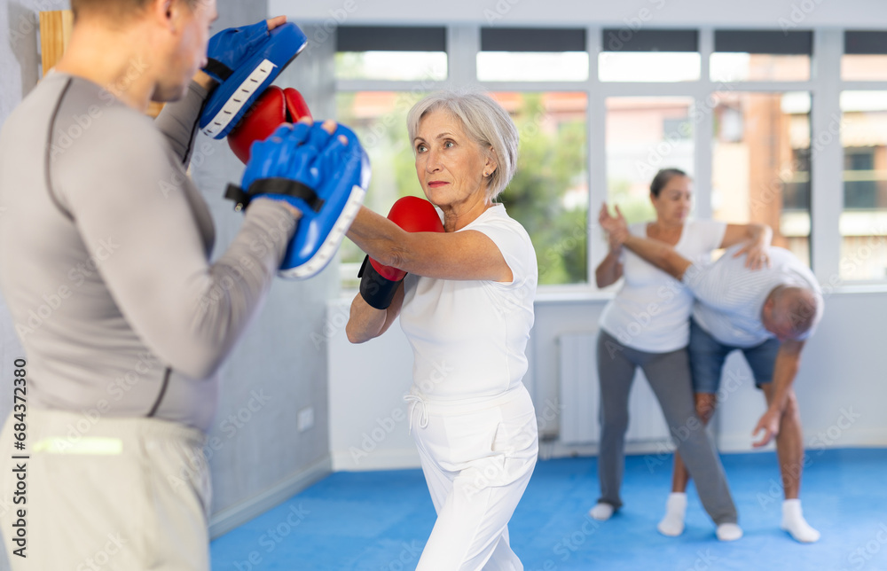 Senior lady and male couch are boxing in gym, man helps student to work ...