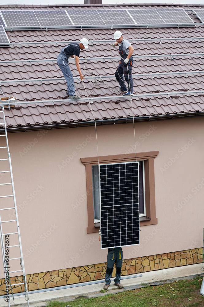 Men technicians lifting up photovoltaic solar moduls on roof of house ...
