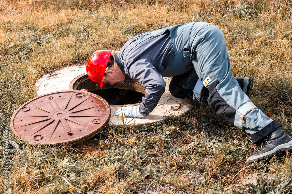 plumber in a hard hat leaned over a water well to check the water meter ...