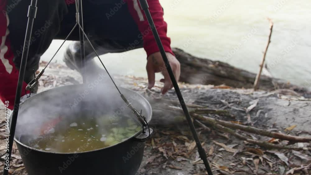 cooking on a hanging cauldron over a fire, on the river bank, a man ...