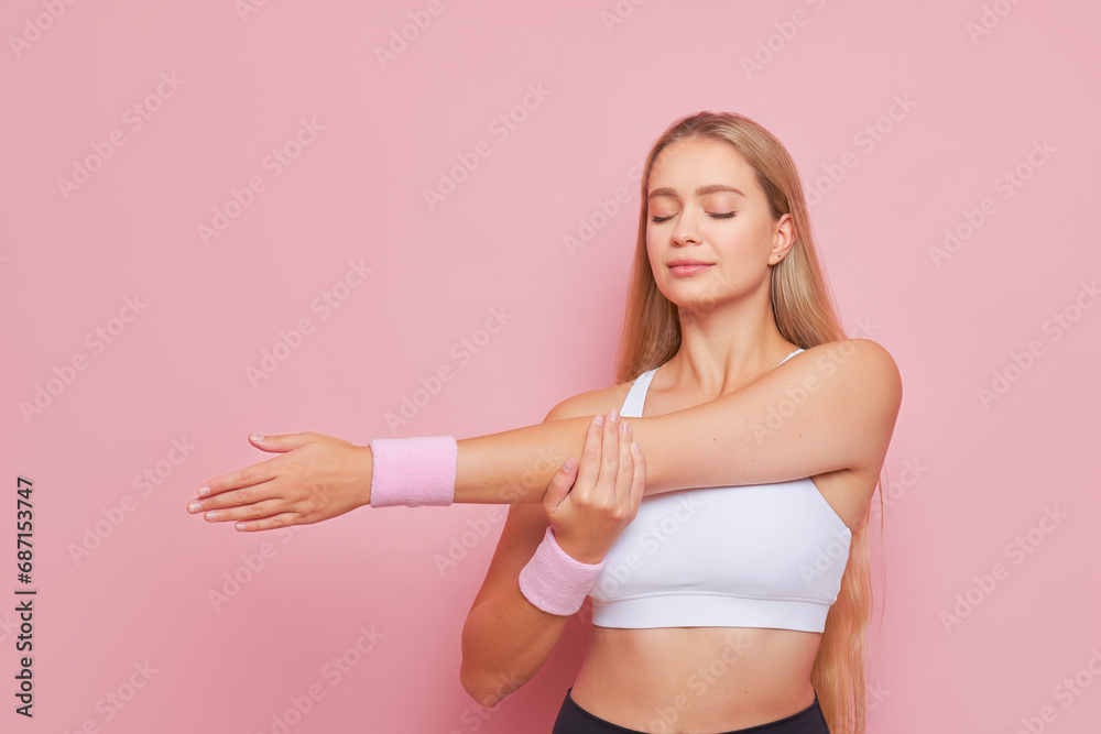 Serious athletic blonde girl in white short sports bustier top doing arm stretching exercises ...