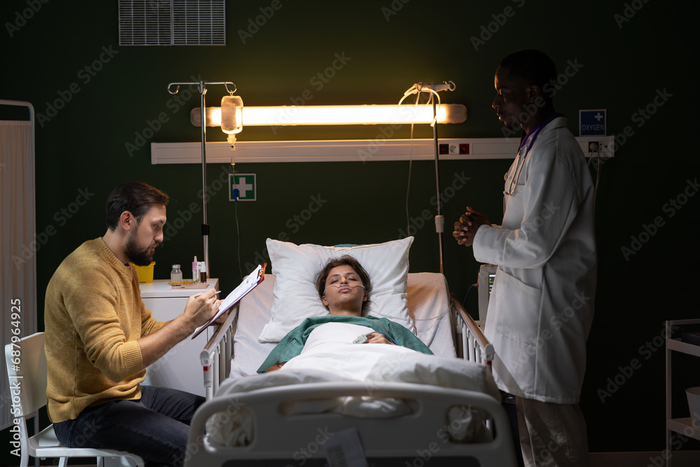 In hospital room, man sits beside bed, attentively examining consent ...