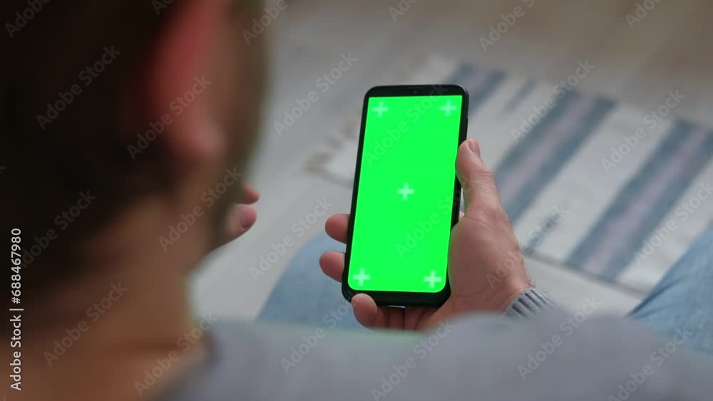 Young man sitting at home holding smartphone green mock-up screen in hand. Male person using ...