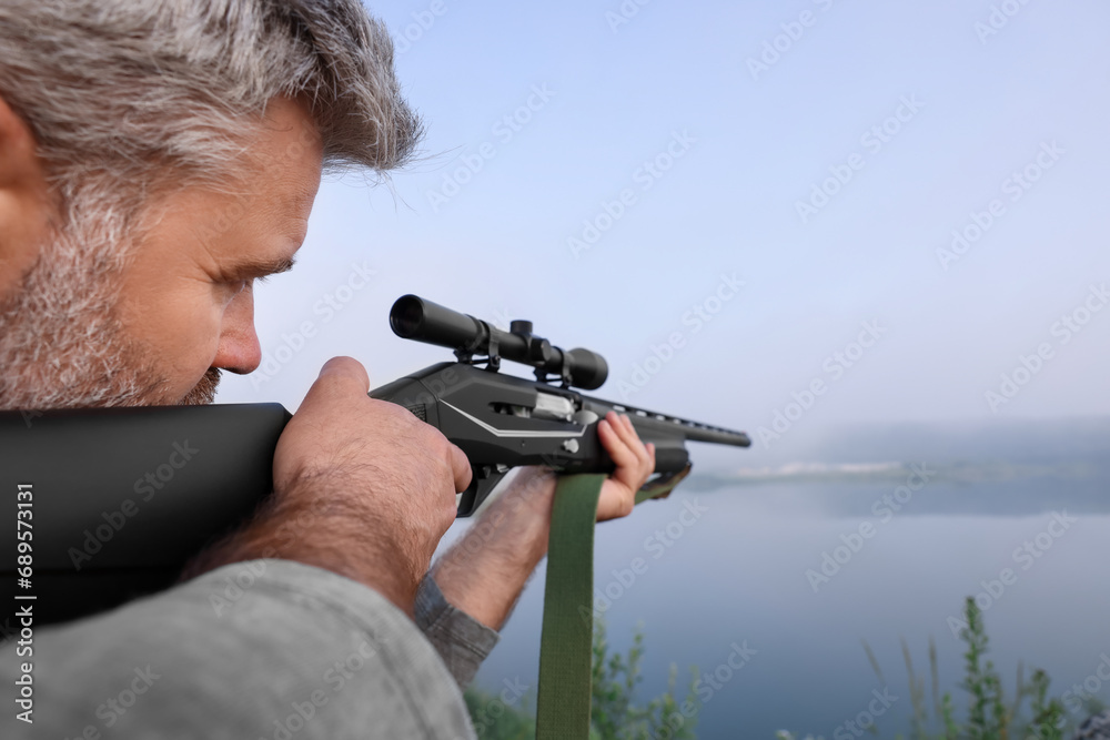 Man aiming with hunting rifle near lake outdoors, closeup. Space for ...