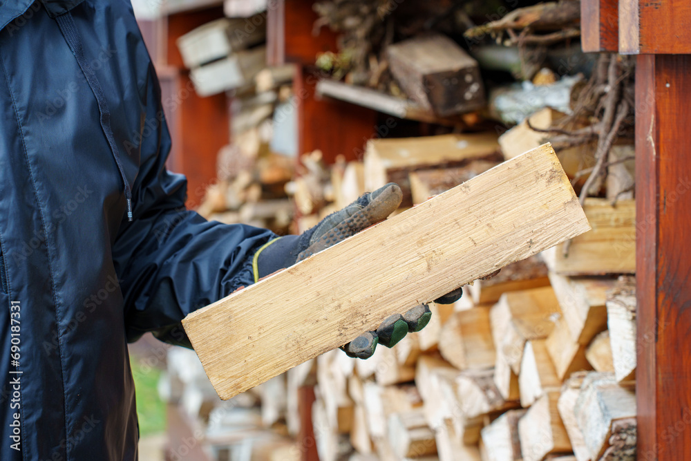 A man in a dark jacket and garden gloves carrying a freshly chopped log ...
