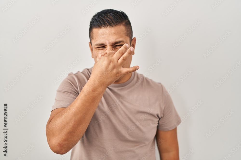 Hispanic young man standing over white background smelling something ...